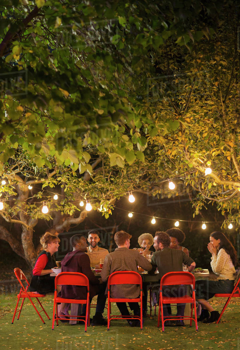 Friends enjoying dinner garden party under trees with fairy lights ...