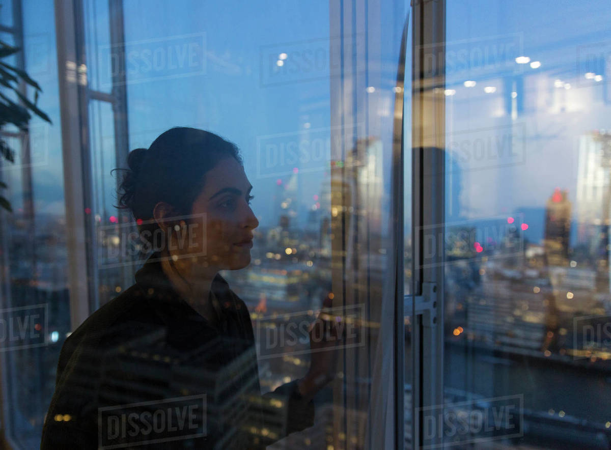 Thoughtful business woman working late at highrise office window ...