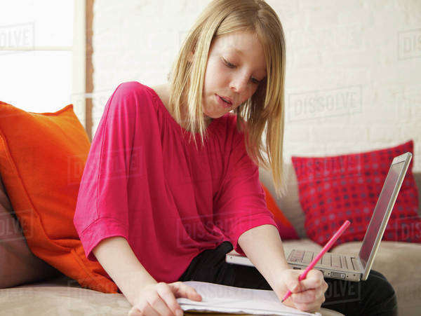 A girl sitting on a sofa with a laptop and writing in a notebook ...
