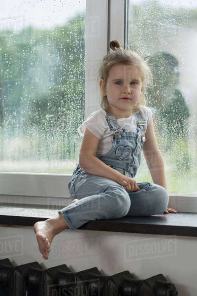 Portrait of cute girl sitting on window sill at home during rainy ...