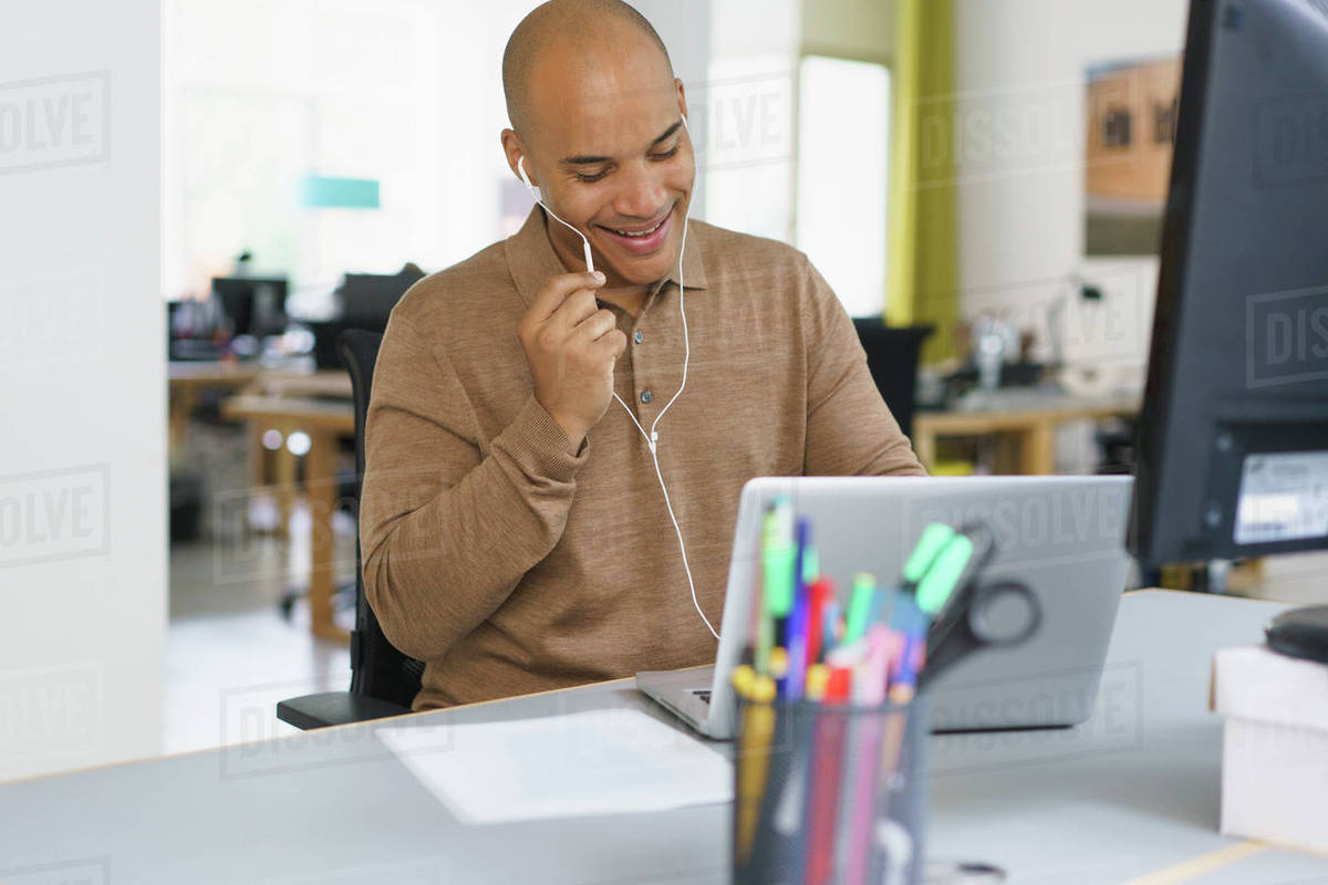 Smiling businessman using headphones while working on laptop at office ...