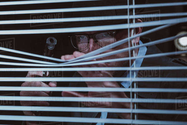 Young man with gun peeking through window blinds - Stock Photo - Dissolve