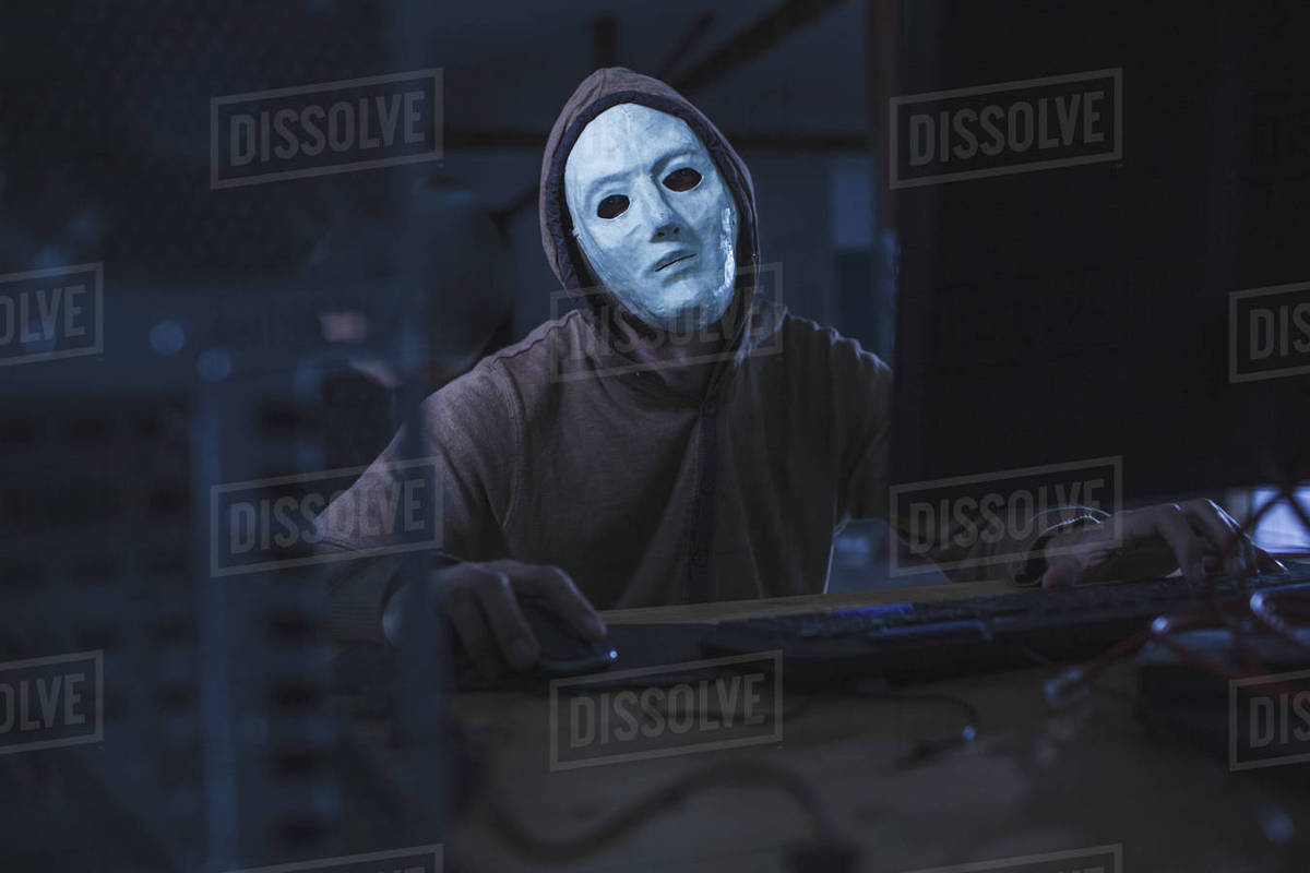 Computer hacker wearing mask and hood using computer while sitting at ...