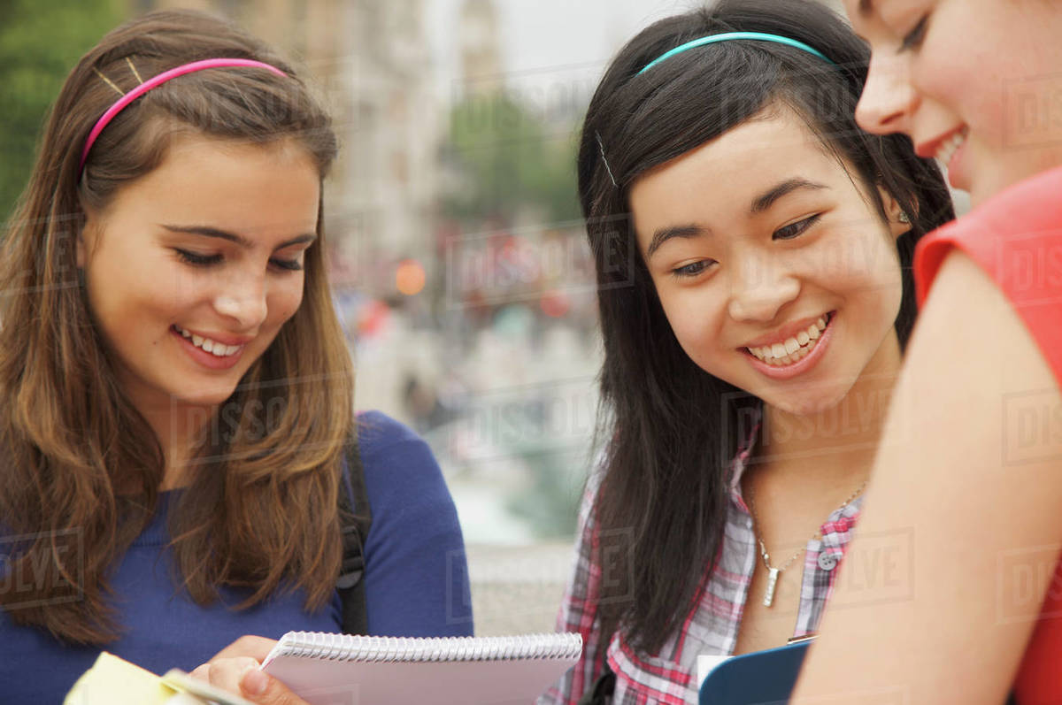 Three smiling teenaged girls reading, writing and holding note pads ...